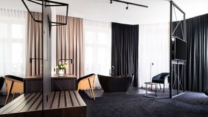 Quarto de hotel moderno com escrivaninha de madeira, duas cadeiras, banheira preta, poltrona preta e TV fixada na parede.