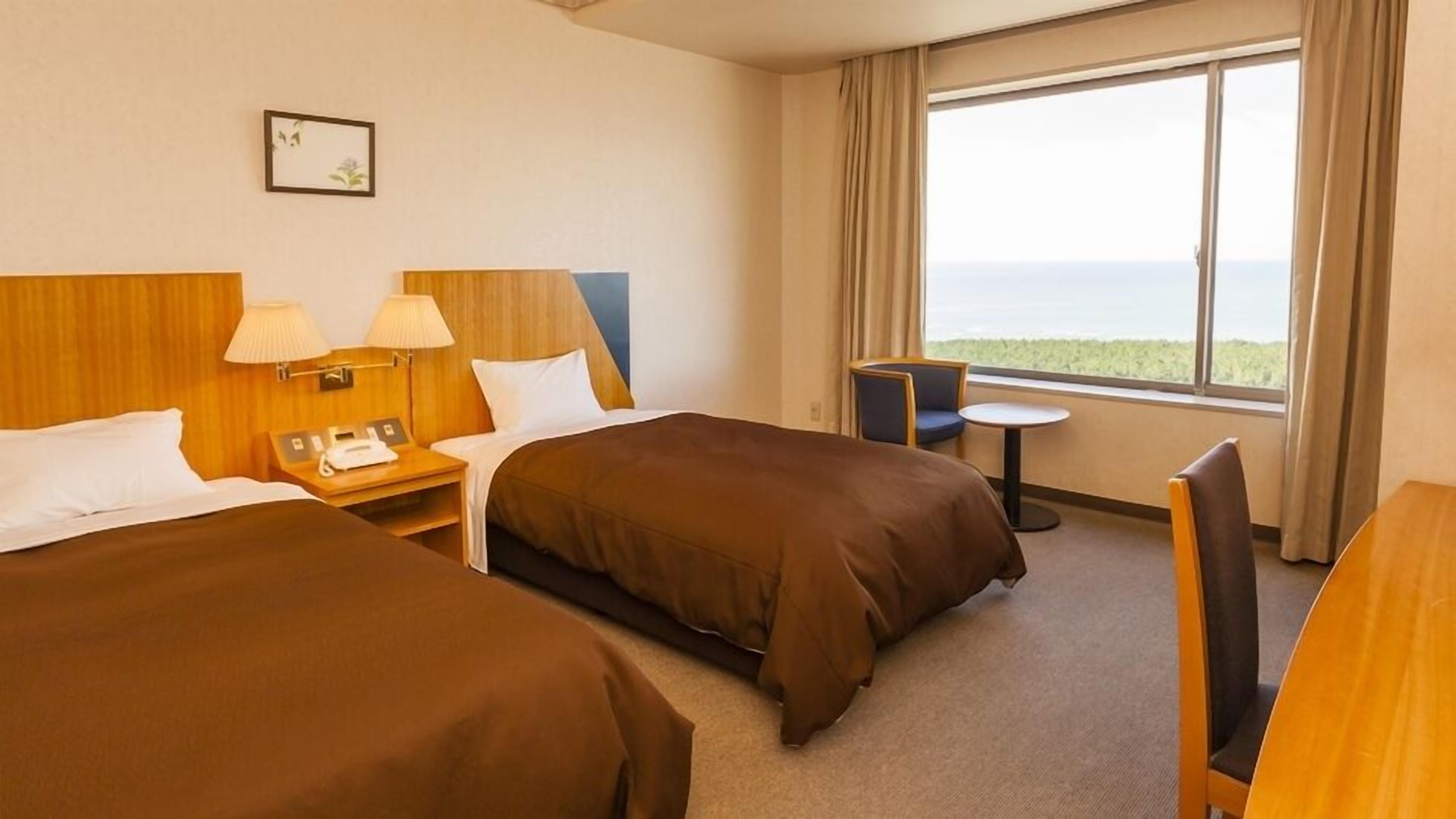 Quarto de hotel com duas camas, uma escrivaninha, uma cadeira, uma mesinha e uma janela com vista.