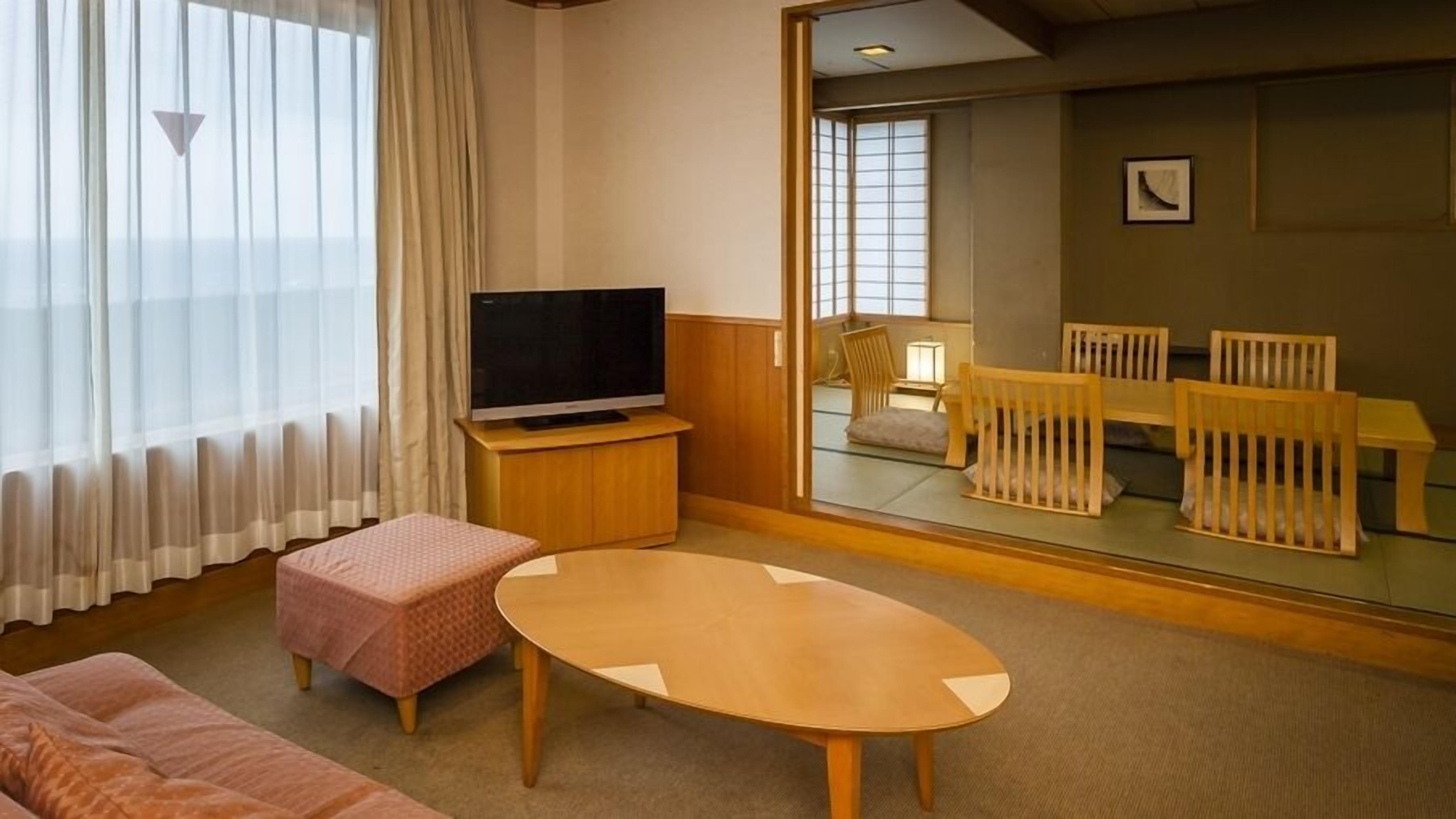 Quarto de hotel com televisão, sofá, mesa pequena e vista para uma área de refeições através de uma divisória de vidro.