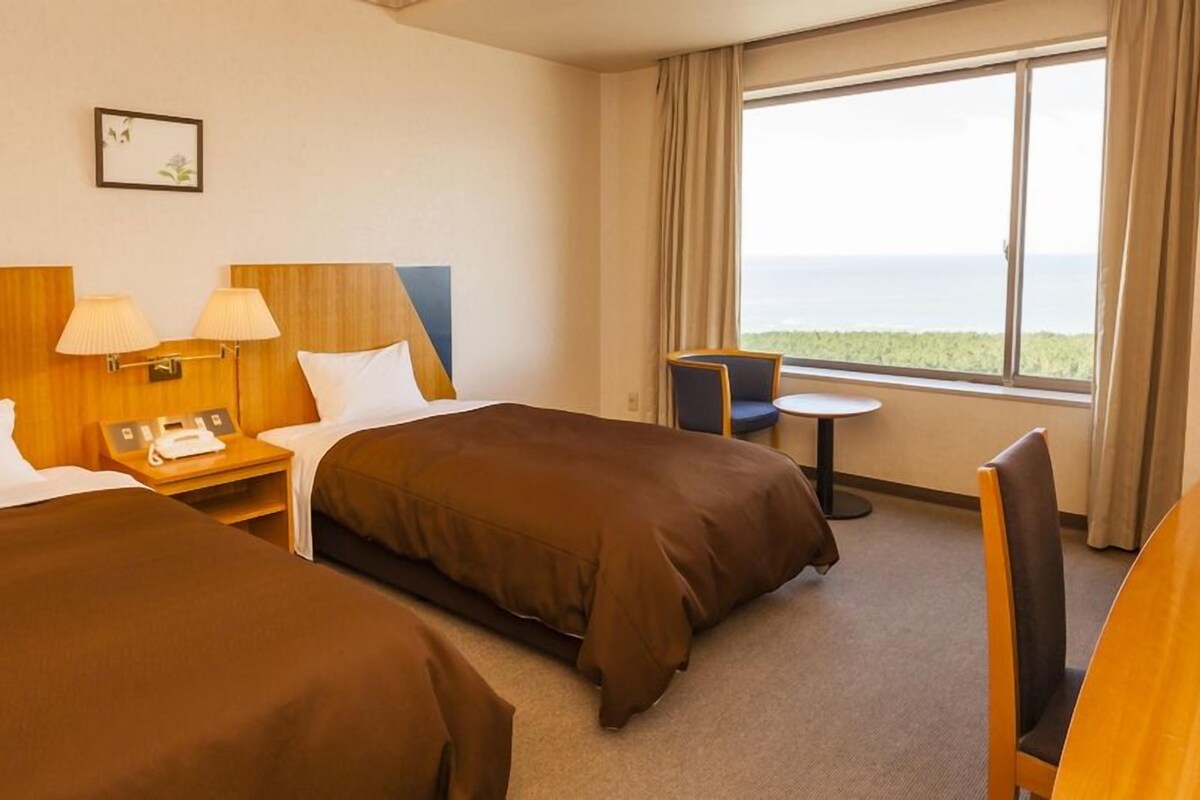 Quarto de hotel com duas camas, uma escrivaninha, uma cadeira, uma mesinha e uma janela com vista.