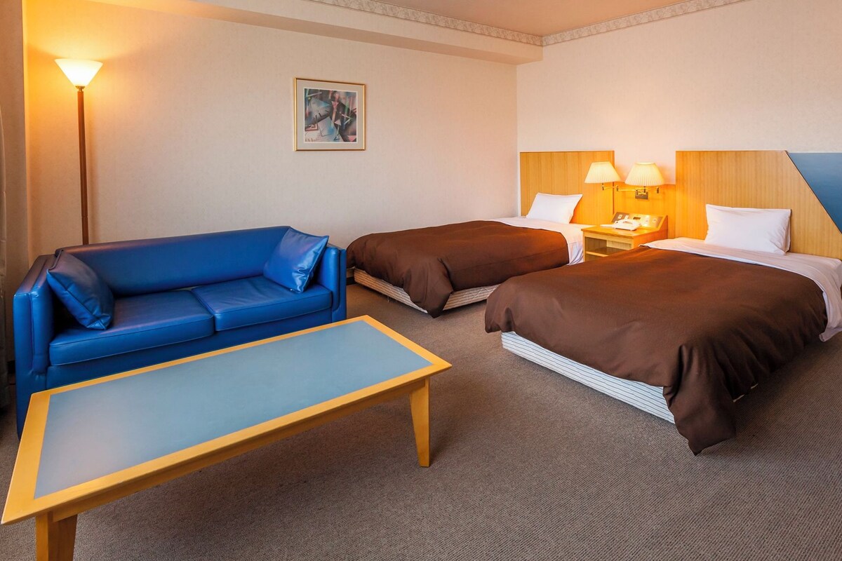 Quarto de hotel com sofá azul, duas camas de solteiro com roupa de cama marrom, mesa de cabeceira de madeira, mesa e abajur.