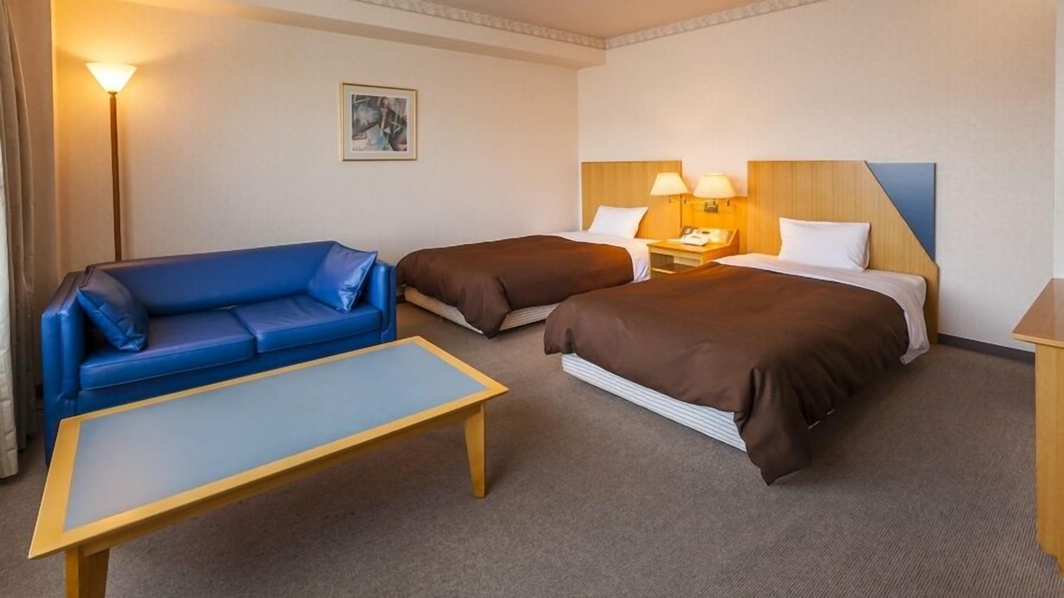 Quarto de hotel com um sofá azul, duas camas de solteiro, uma mesinha de cabeceira de madeira, um abajur e um quadro na parede.