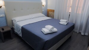 Uma cama bem arrumada com colcha azul e lençóis brancos, uma mesinha de cabeceira com controle remoto e uma luminária fixada na parede.