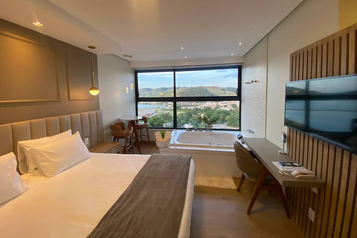 Quarto de hotel com cama, escrivaninha, cadeira, televisão e uma grande janela com vista para a cidade.