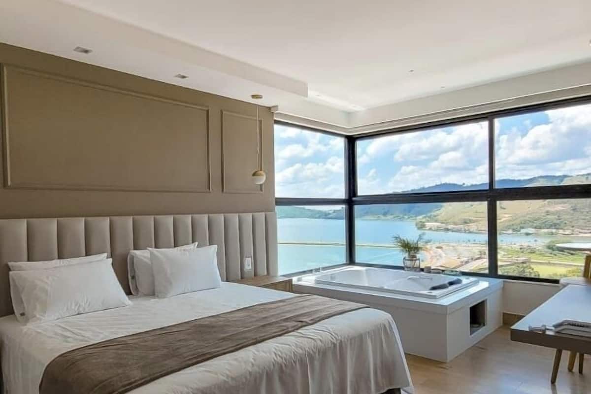 Quarto moderno com uma cama grande, vista para um lago e um pequeno espaço de estar.