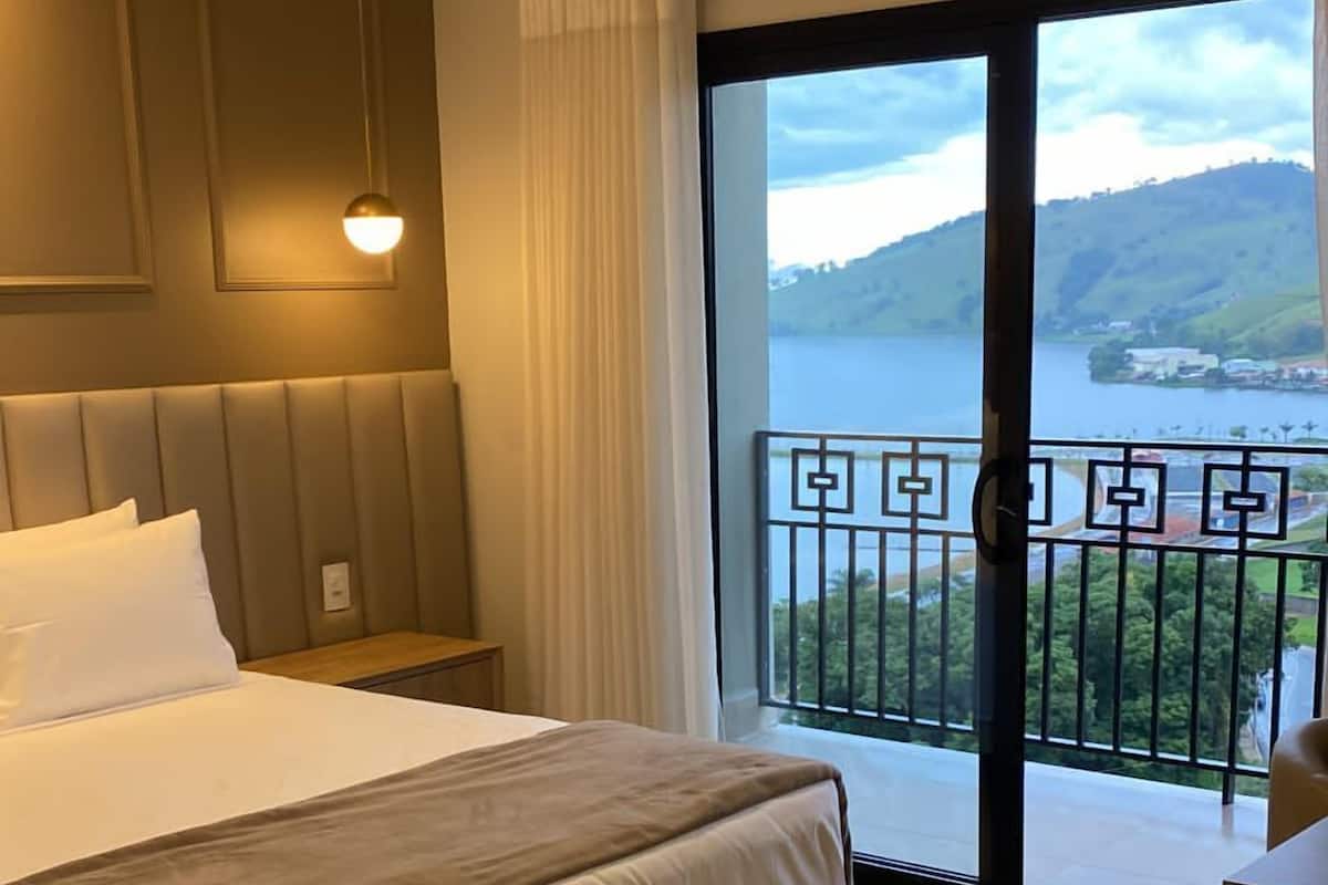 Quarto de hotel com uma cama grande, uma mesinha de cabeceira, uma cadeira e uma varanda com vista para um lago e colinas.