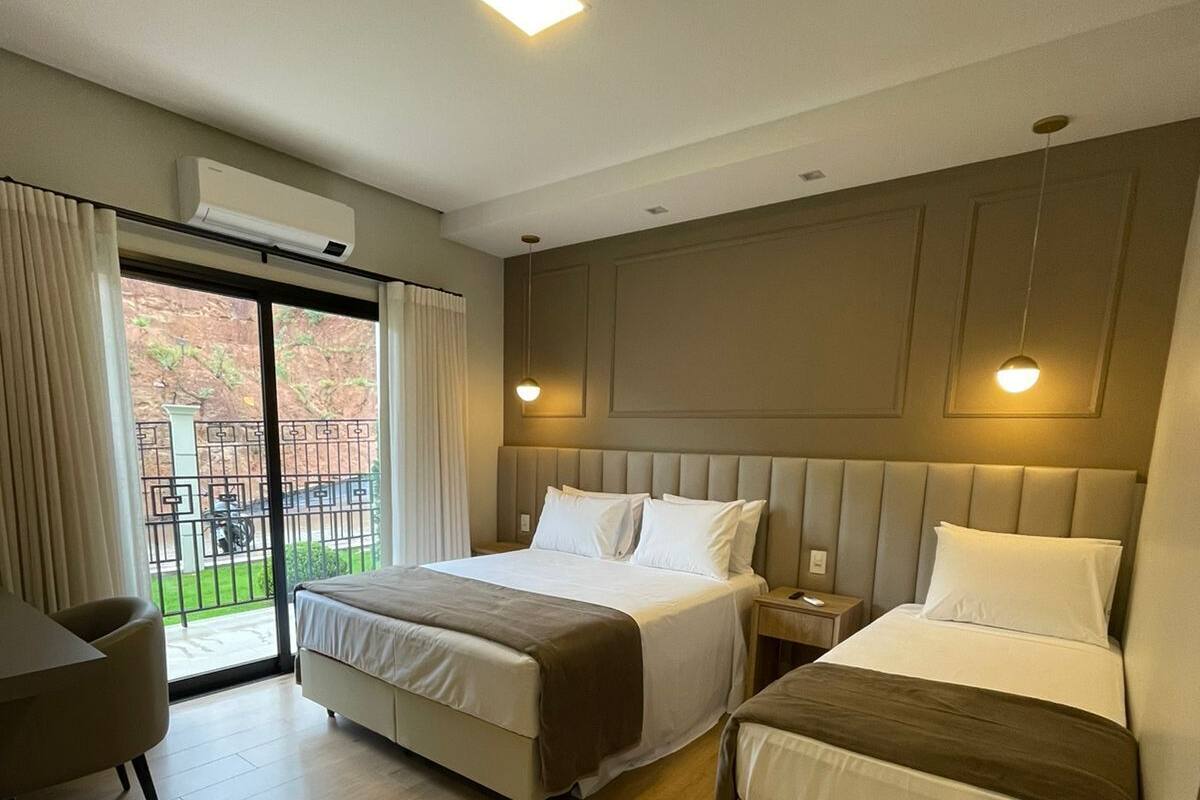 Quarto de hotel com duas camas, uma mesa de trabalho e porta de vidro que se abre para uma área externa.