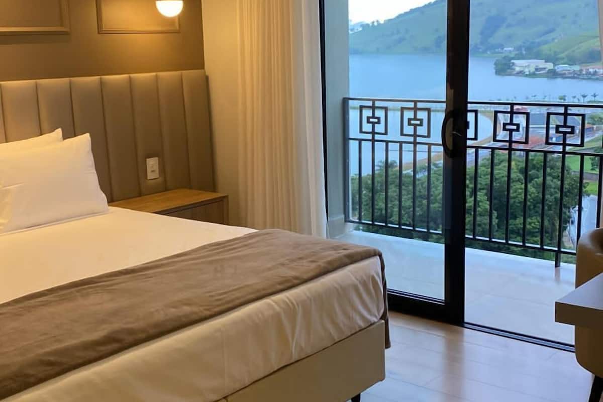Quarto com cama grande, mesa de cabeceira, cadeira e varanda com vista para um lago.