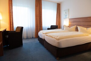 Quarto de hotel com uma cama grande, duas cadeiras, uma escrivaninha e um abajur.
