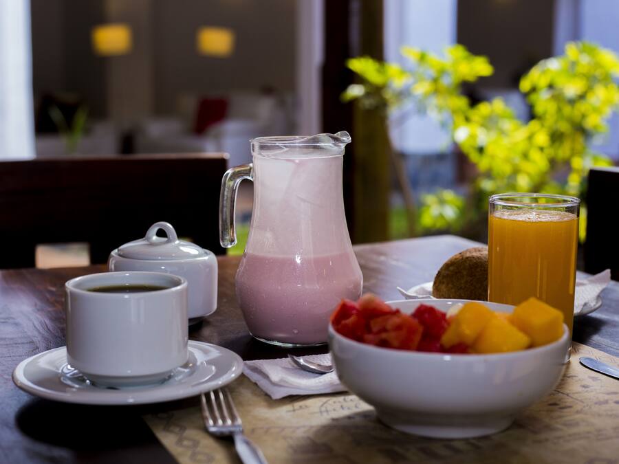 Uma mesa de madeira com um jarro de smoothie rosa, um copo de suco de laranja, uma tigela de frutas e uma xícara de café.