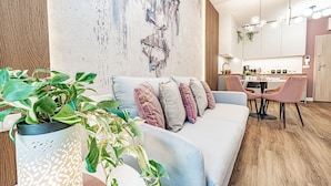 Uma sala de estar moderna com um sofá cinza claro, vários almofados e uma planta verde grande. Uma área de cozinha com mesa e cadeiras é visível ao fundo.