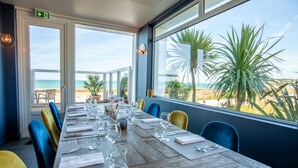 Uma sala de jantar com mesa de madeira posta para seis, grandes janelas com vista para a praia e cadeiras coloridas.