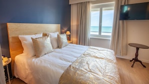 Quarto de hotel com uma cama grande, televisão na parede, mesa de cabeceira e vista para o mar pela janela.