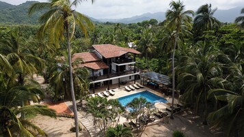 Uma casa de vários andares com piscina, rodeada por palmeiras e um cenário montanhoso.
