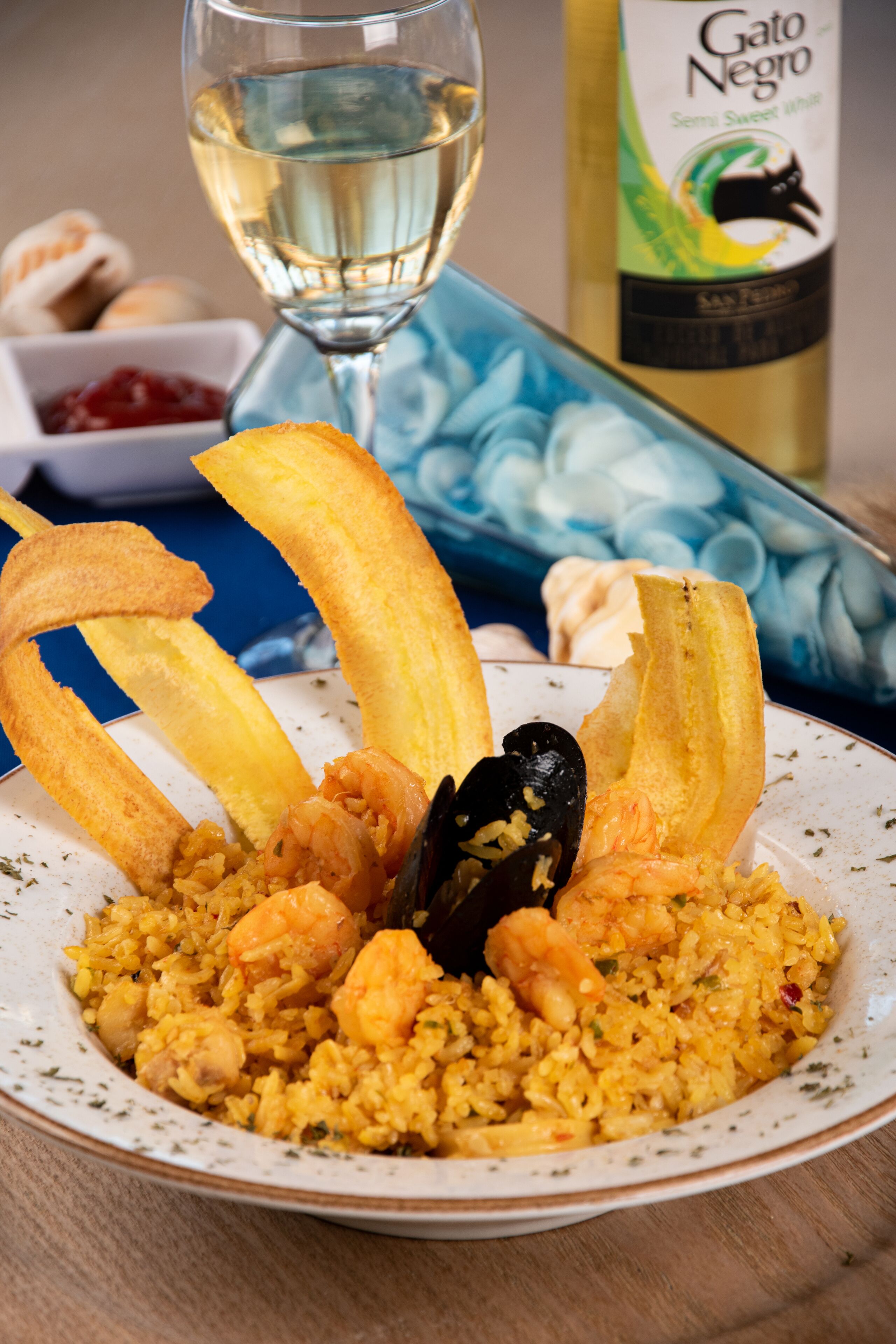 Prato de arroz com camarões e mexilhões, acompanhado por um copo de vinho branco e uma garrafa de vinho Gato Negro.