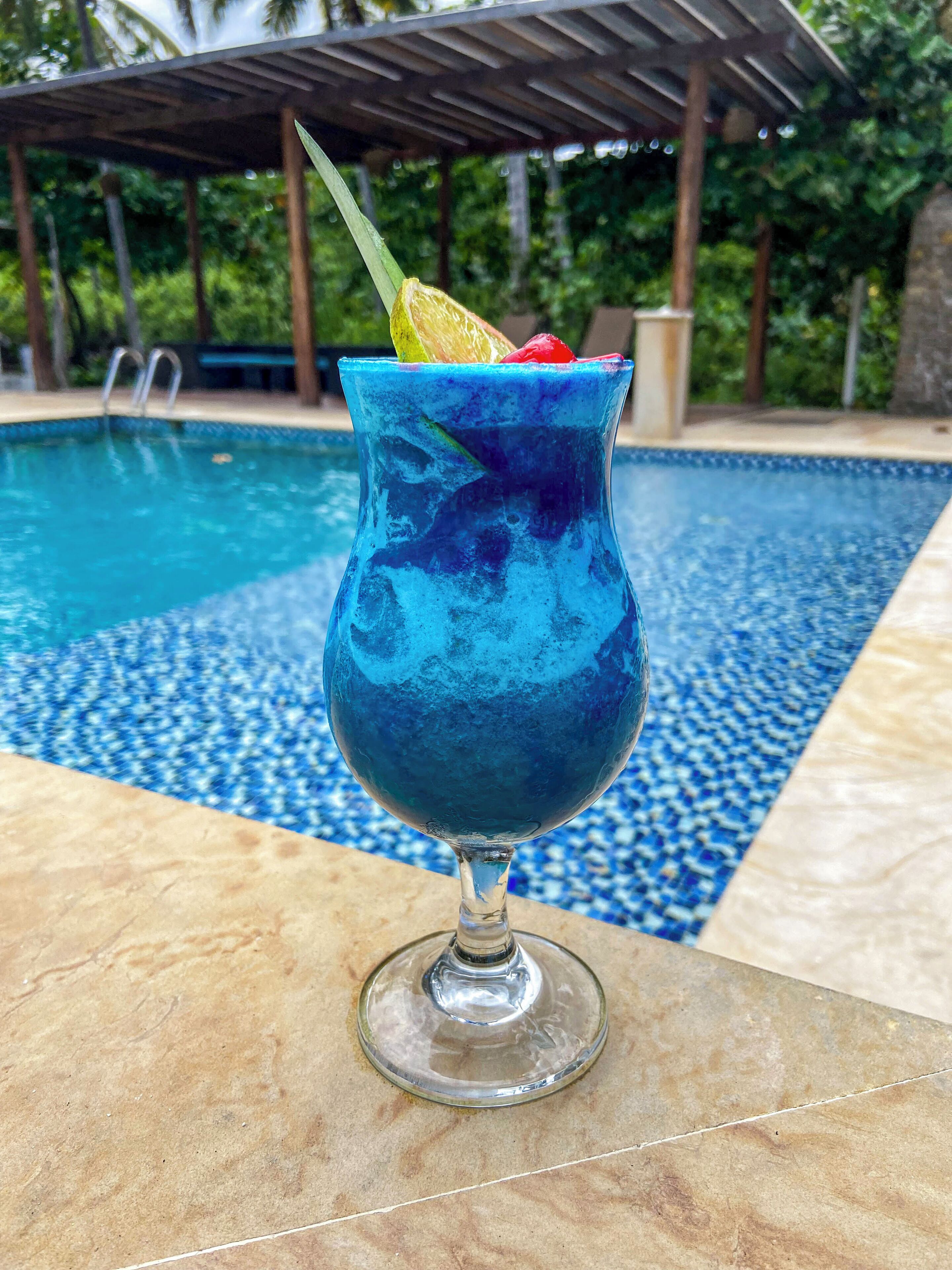 Um cocktail azul num copo com uma fatia de limão e uma cereja, sobre uma superfície azulejada perto de uma piscina.