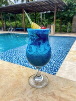 Um cocktail azul num copo com uma fatia de limão e uma cereja, sobre uma superfície azulejada perto de uma piscina.
