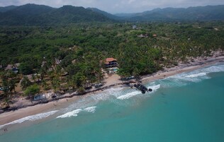 Vista aérea de uma praia com uma casa rodeada por palmeiras, areia dourada e águas azul-turquesa.