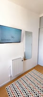 Quarto com radiador branco, tapete estampado, televisão exibindo uma cena de praia e porta com espelho.
