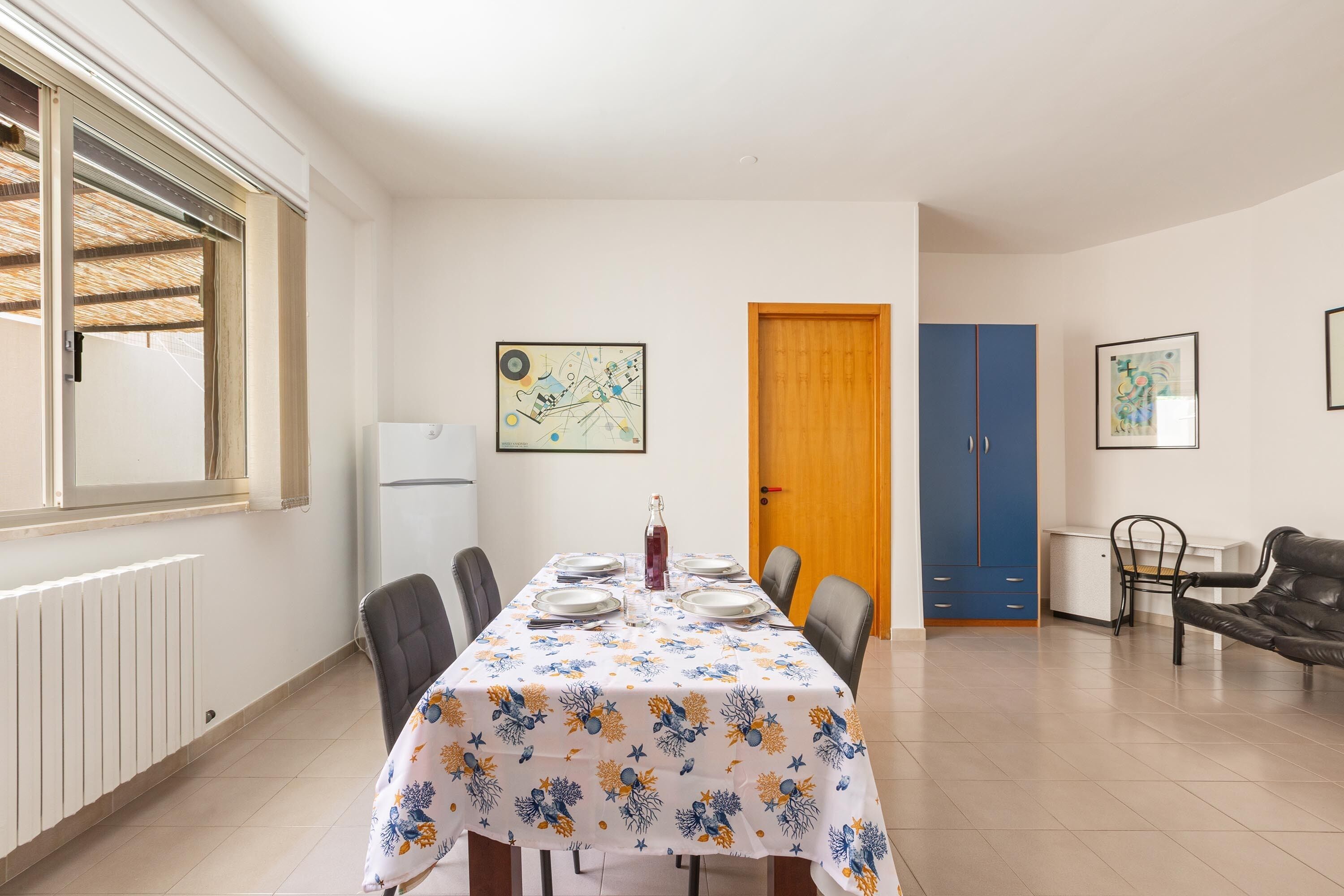 Uma sala de jantar com mesa posta para quatro pessoas, um frigorífico, um guarda-roupa azul e uma janela com persianas.