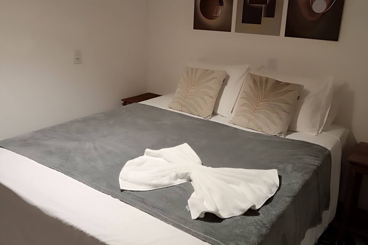 Cama com colcha cinza, almofadas brancas e manta branca. Três quadros decoram a parede acima da cama.
