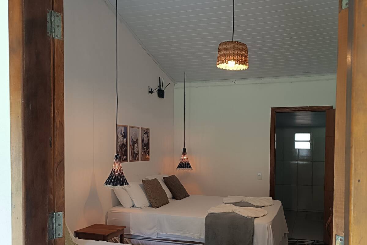 Quarto com duas camas, uma mesinha, luminárias na parede e uma porta que dá para outro cômodo.