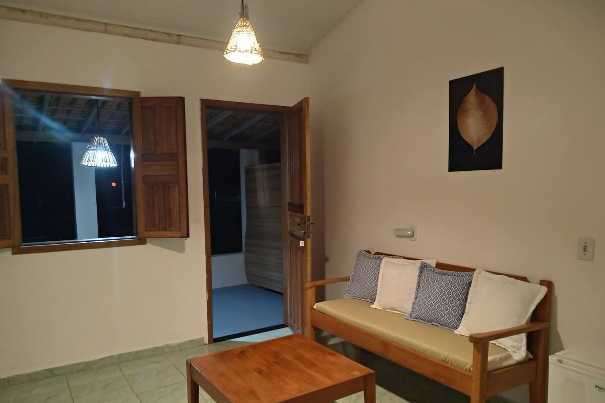 Quarto com sofá de madeira, mesa de madeira, luminária pendente, janela com persianas de madeira e porta que dá para outro cômodo.