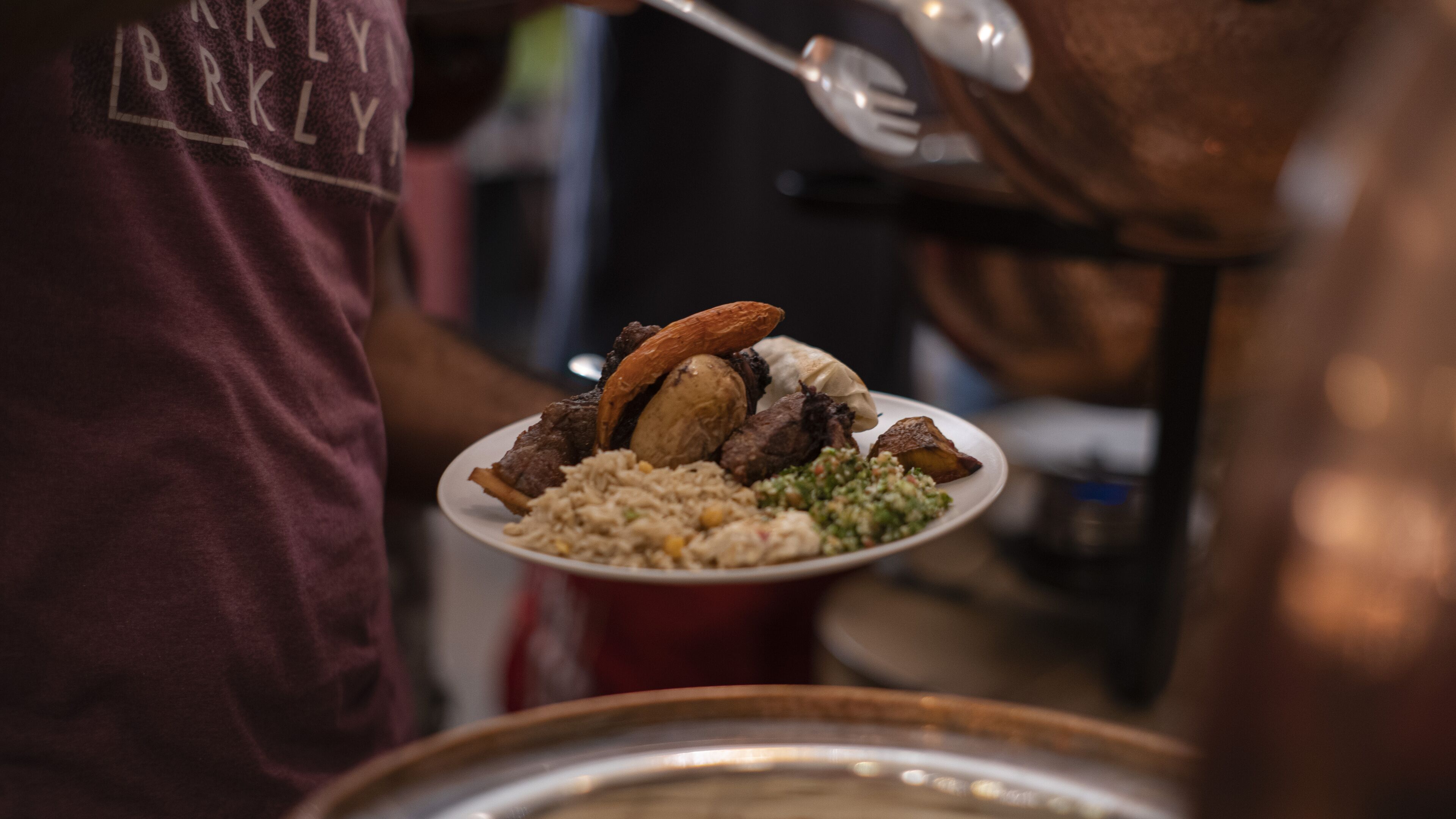 Uma pessoa servindo um prato de comida com diversos itens, incluindo carne, batatas e arroz.