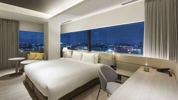 Quarto de hotel moderno com uma cama grande, uma escrivaninha e uma cadeira. Janelas amplas oferecem vista para o horizonte da cidade.