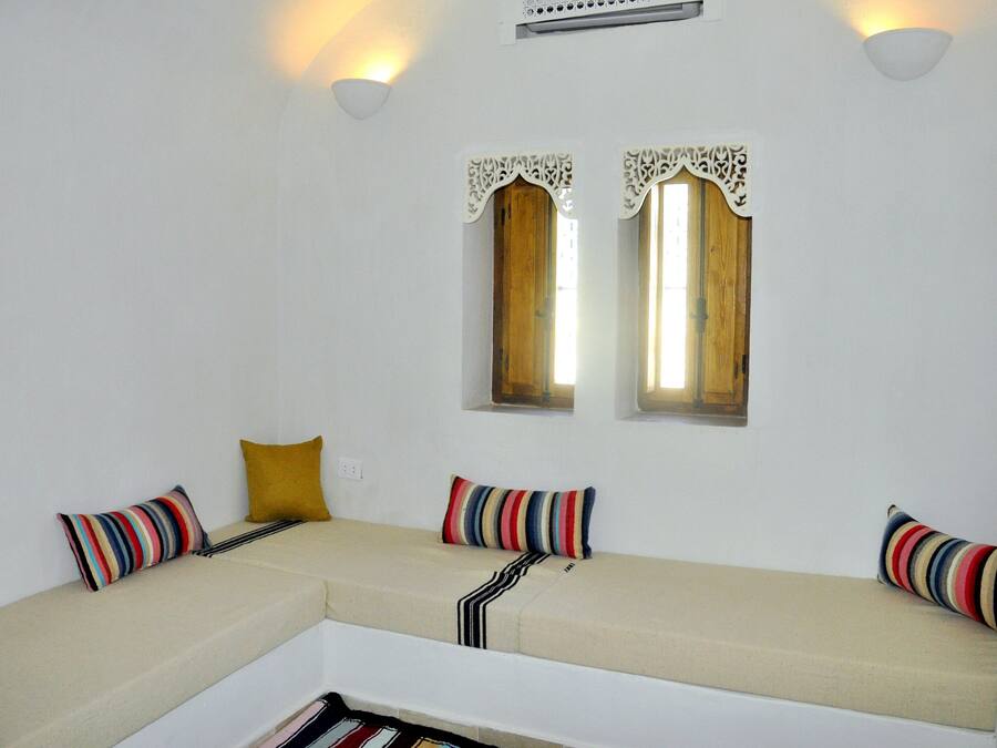 Quarto com sofá bege, duas janelas com molduras de madeira e detalhe rendado branco.