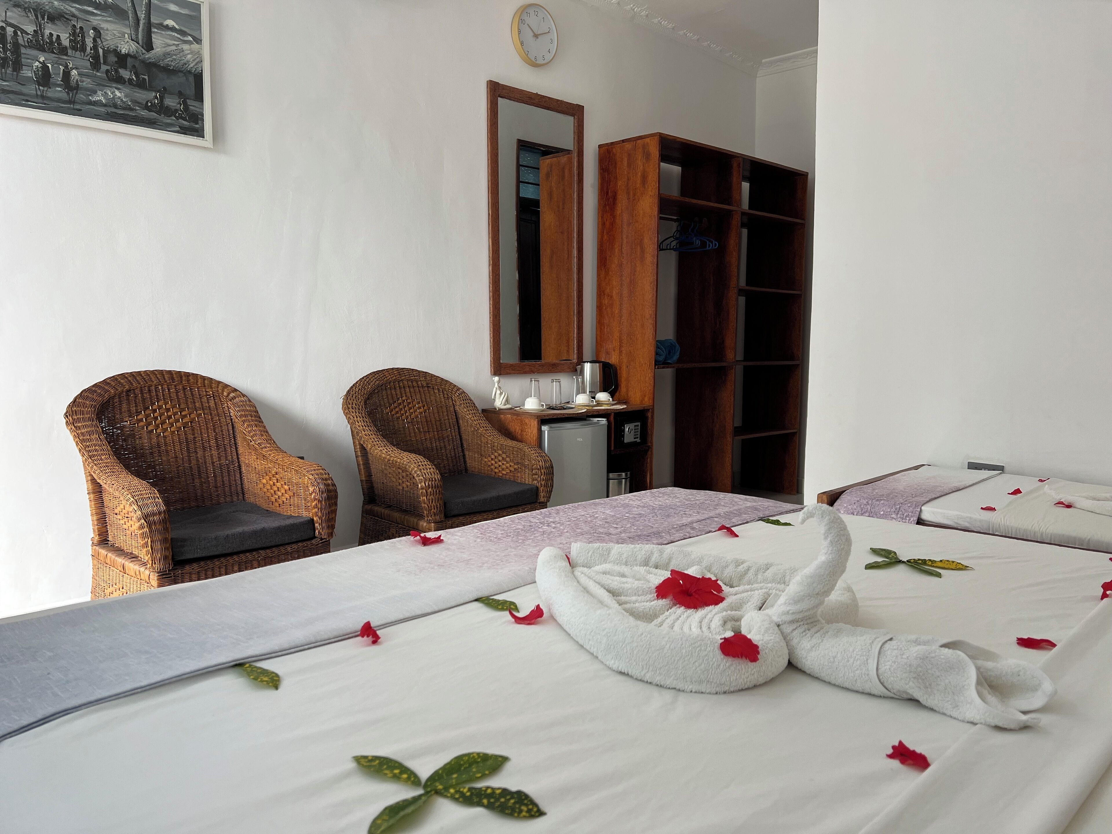 Quarto de hotel com duas cadeiras de vime, uma cama com edredom branco e flores vermelhas, um guarda-roupa de madeira e uma pequena mesa com chaleira e chávenas.