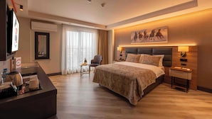 Quarto de hotel com uma cama grande, uma escrivaninha com cafeteira, uma cadeira, uma TV e um quadro na parede.