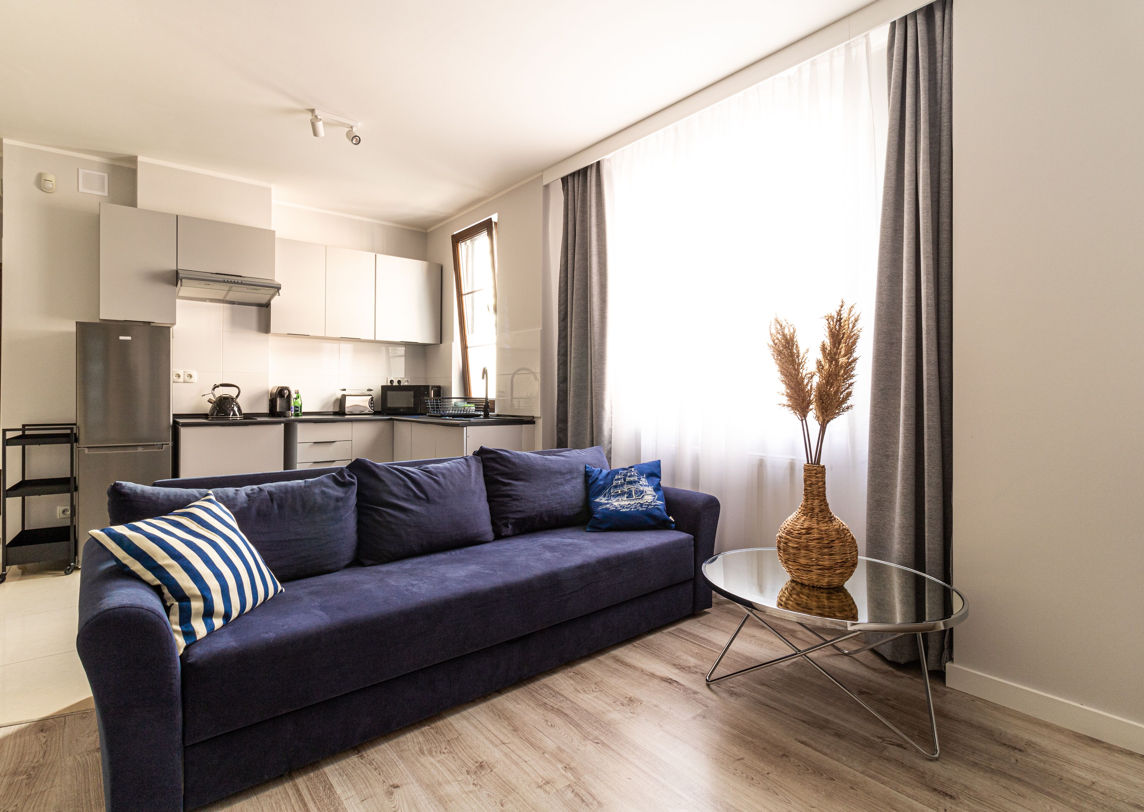 Uma sala de estar moderna com um sofá azul escuro, uma mesa de centro de vidro e uma cozinha ao fundo.