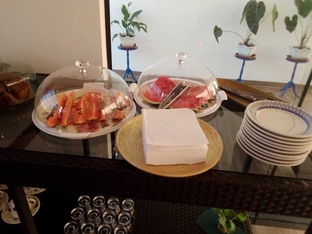 Disposição de buffet com pratos de comida cobertos, uma pilha de pratos e uma pequena planta sobre uma mesa.