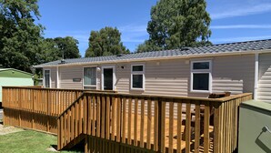 Uma casa móvel com um deck de madeira, um pequeno alpendre e um depósito utilitário verde.