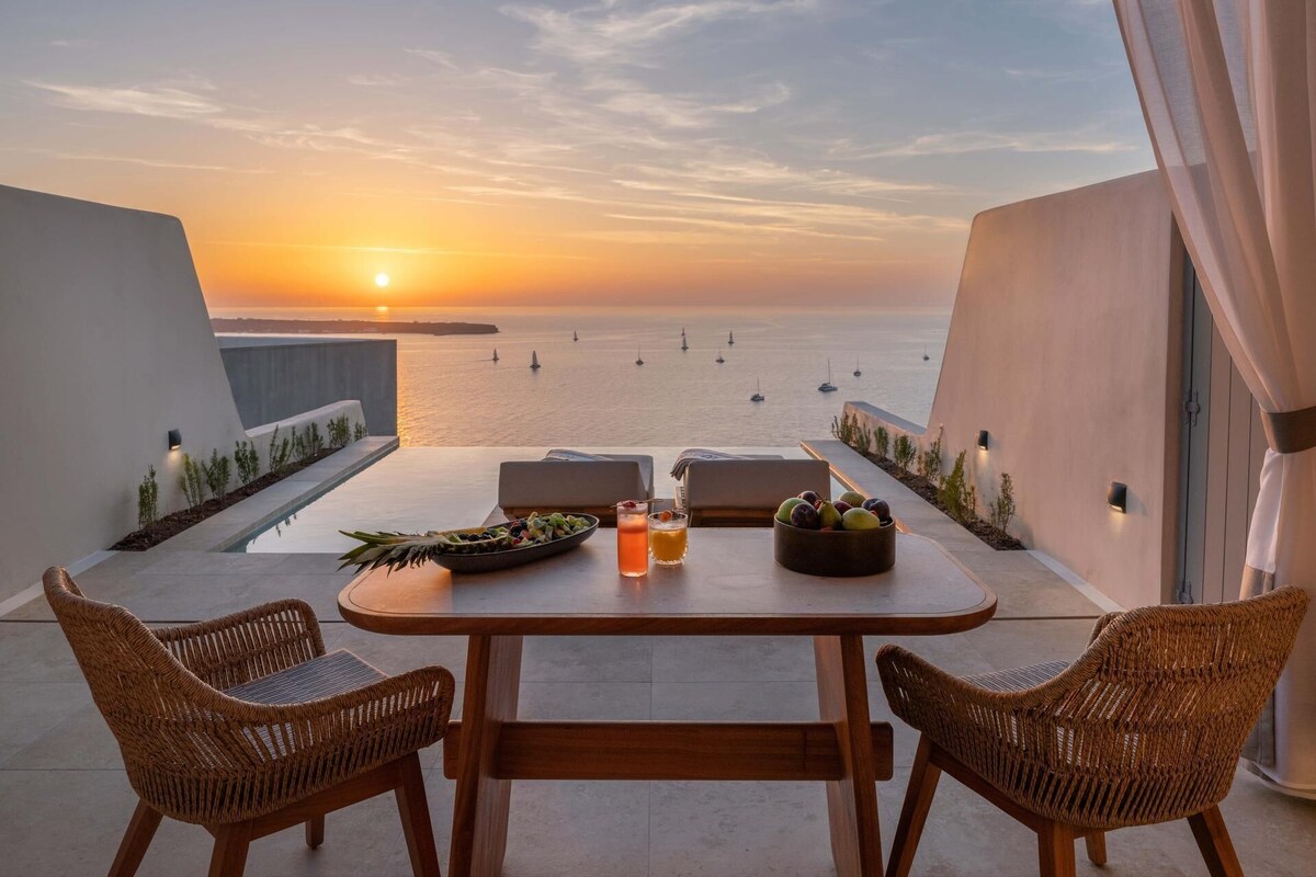 Terraço com mesa posta para dois, com vista para o mar, barcos e um pôr do sol.