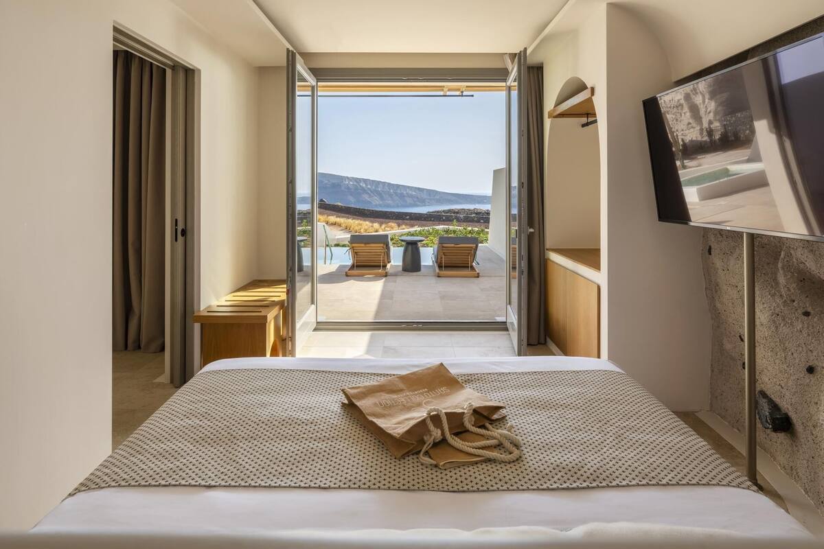 Quarto de hotel com cama, televisão e vista para um pátio com mobiliário exterior e uma montanha ao longe.