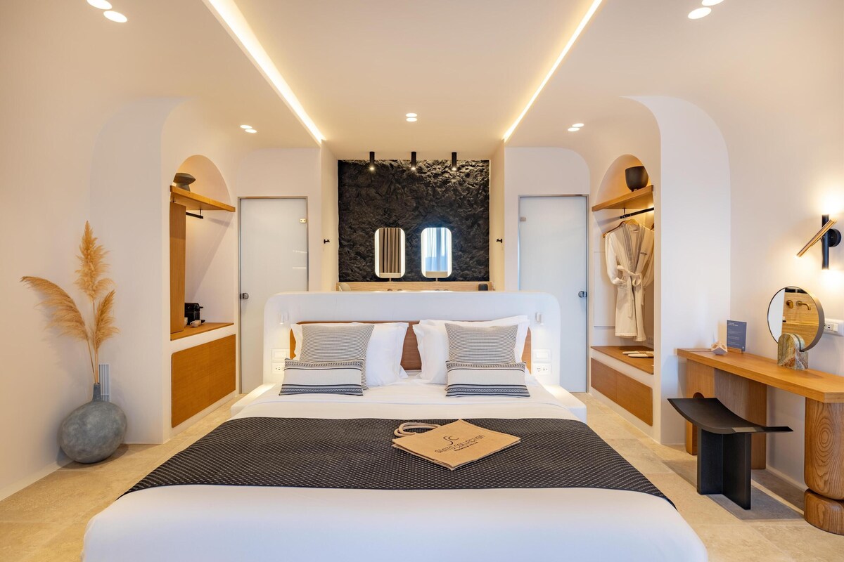Quarto de hotel moderno com uma cama grande, uma mesa de toucador com espelho e um roupão pendurado na parede.