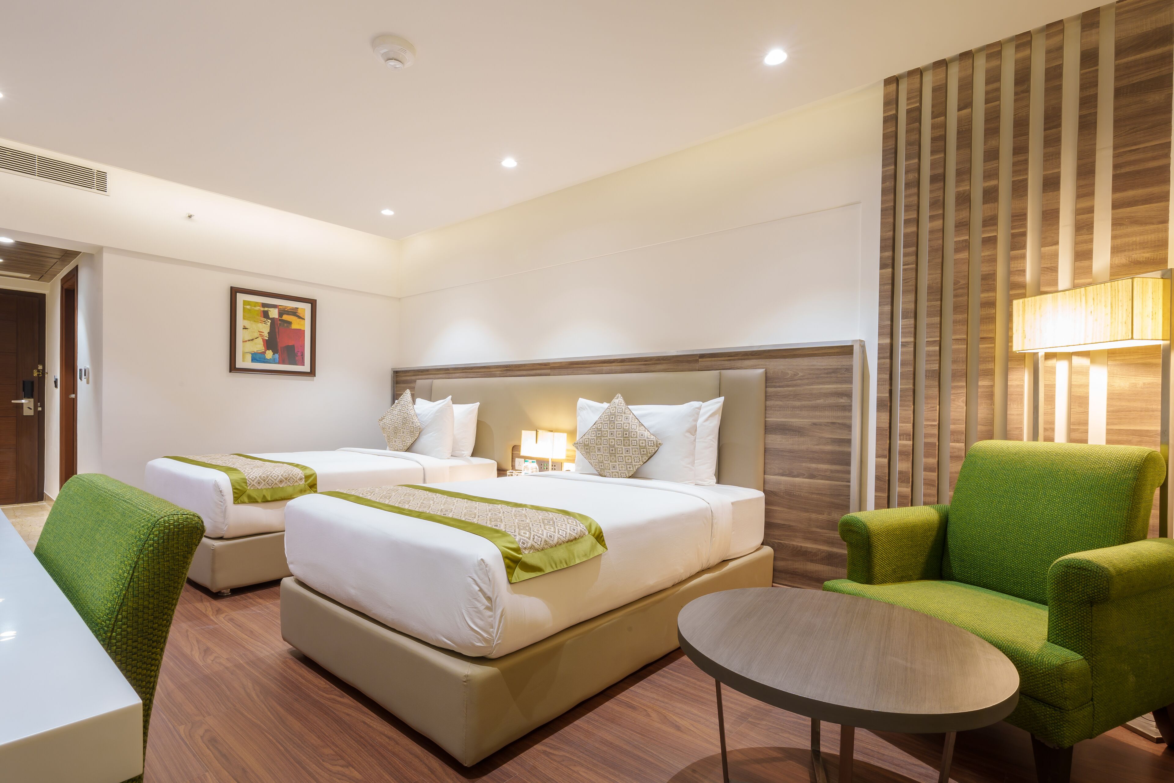Quarto de hotel com duas camas, uma poltrona verde, uma mesinha e um quadro na parede.
