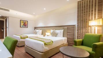 Quarto de hotel com duas camas, uma poltrona verde, uma mesinha e um quadro na parede.