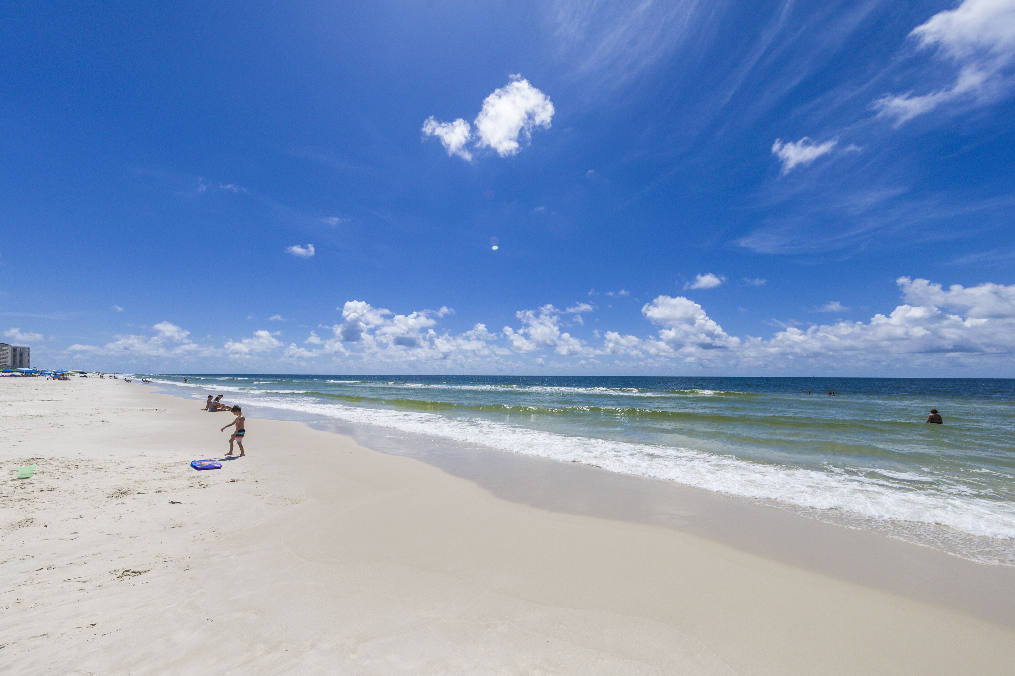 Uma praia com céu claro, ondas calmas e algumas pessoas.