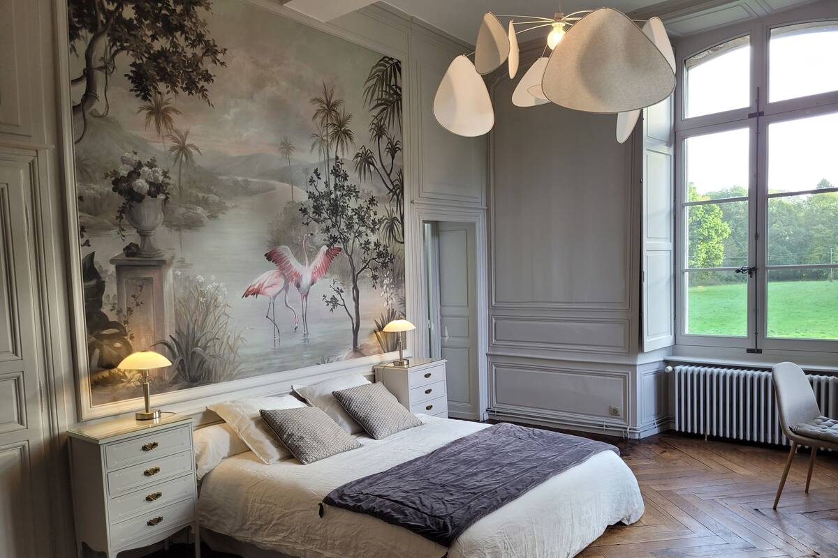 Quarto com um mural grande, uma cama com várias almofadas, mesas de cabeceira com abajures e vista através de uma janela ampla.