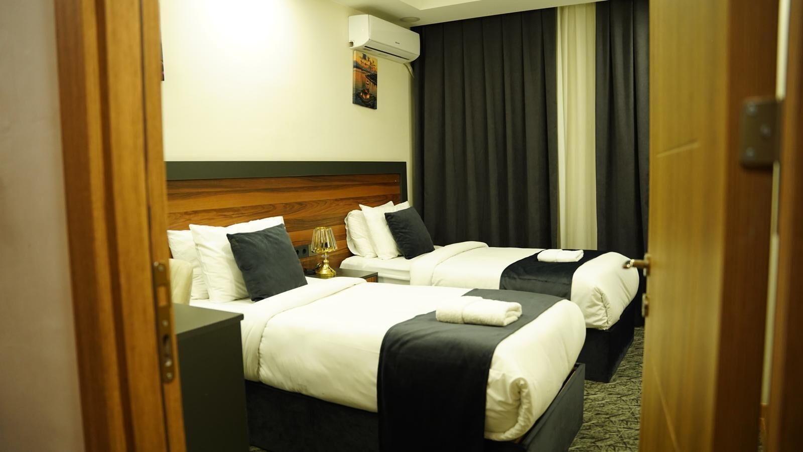 Quarto de hotel com duas camas, cabeceira de madeira, mesa de cabeceira com abajur e uma janela grande com cortinas.