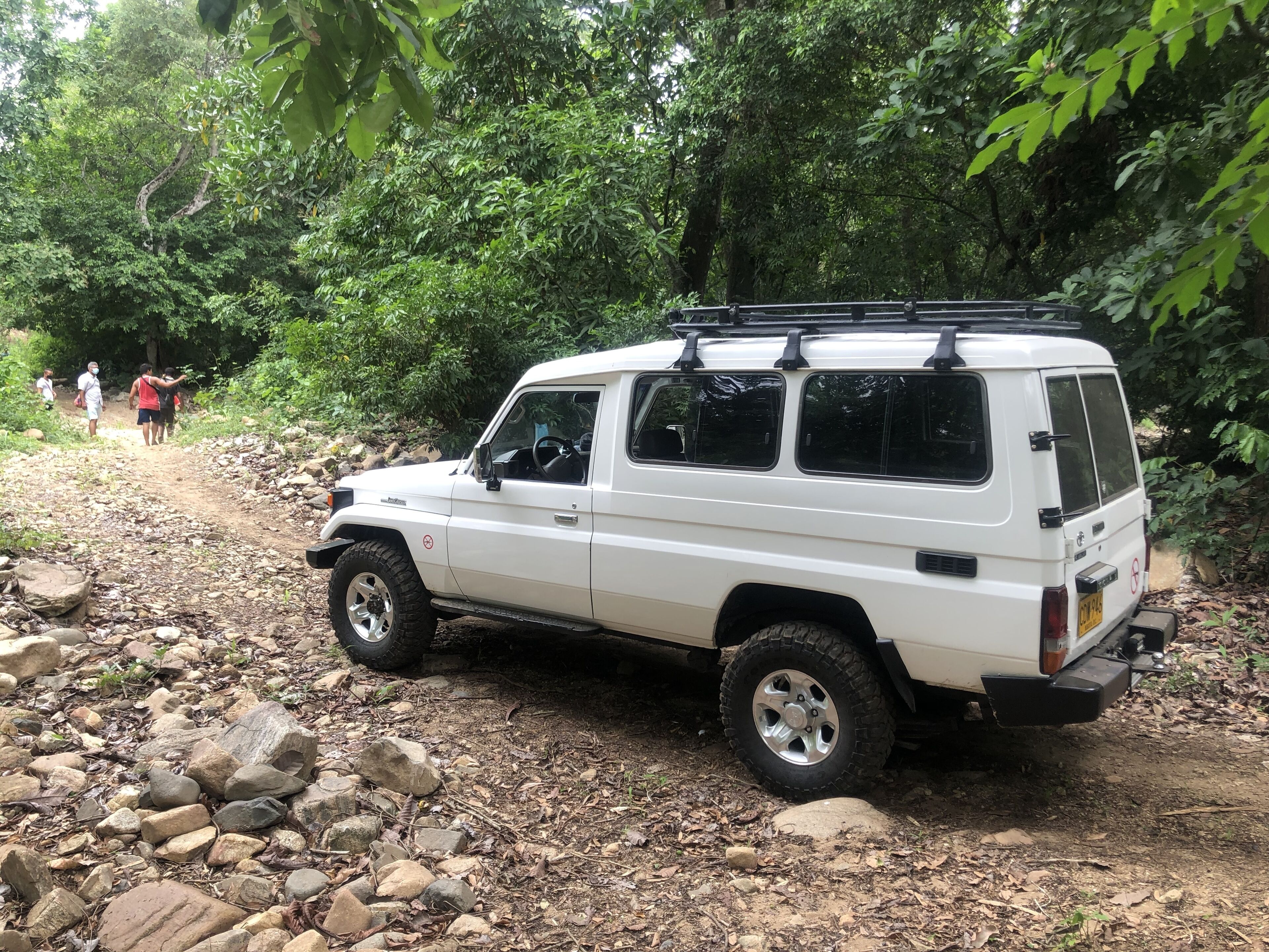 Um SUV branco estacionado numa trilha pedregosa, rodeado por vegetação densa.