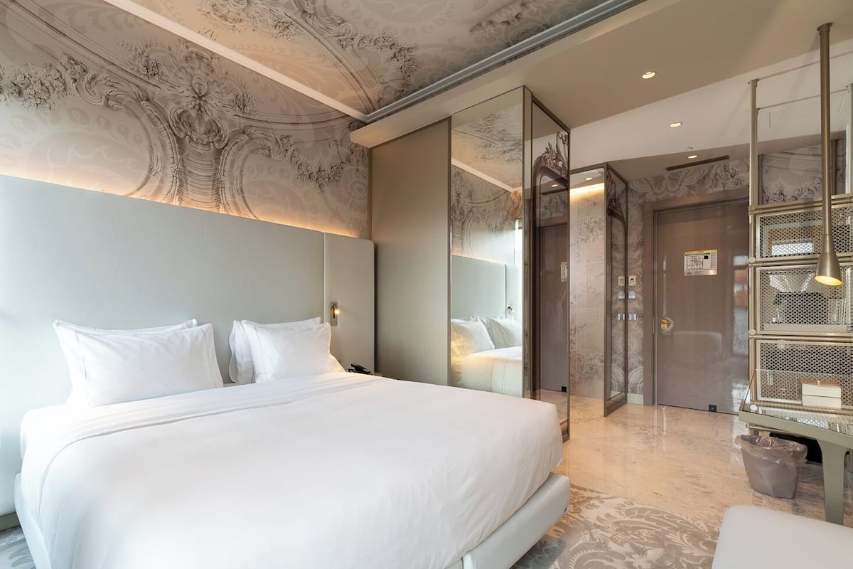 Quarto de hotel moderno com uma cama grande, mesa de cabeceira, espelho e elevador envidraçado.