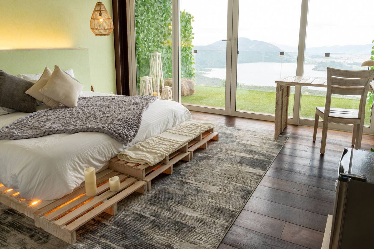 Quarto com cama grande, mesas de cabeceira, vista para um lago e uma cadeira de madeira.