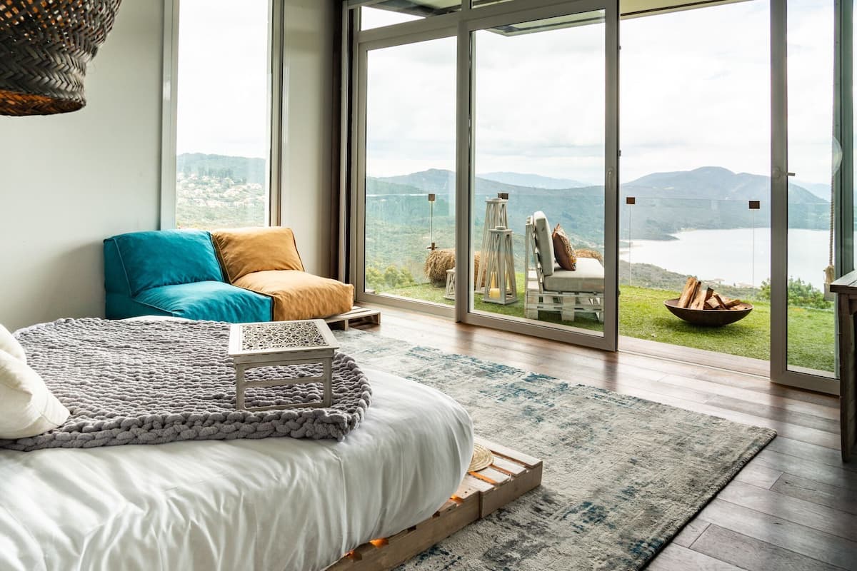 Quarto com uma janela grande, uma cama, uma cadeira azul e vista para um lago e montanhas.