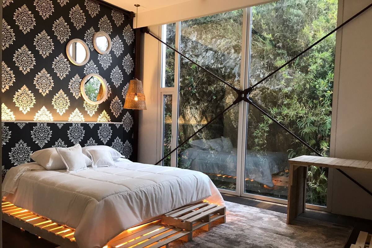 Quarto moderno com uma cama grande, um banco de madeira e vista para o verde através de amplas janelas.