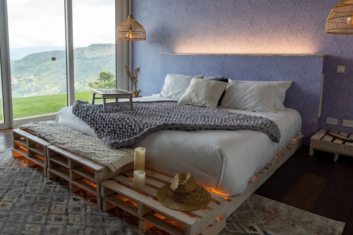 Quarto com cama, um banco de paletes de madeira, mesa de cabeceira e vista para o exterior.