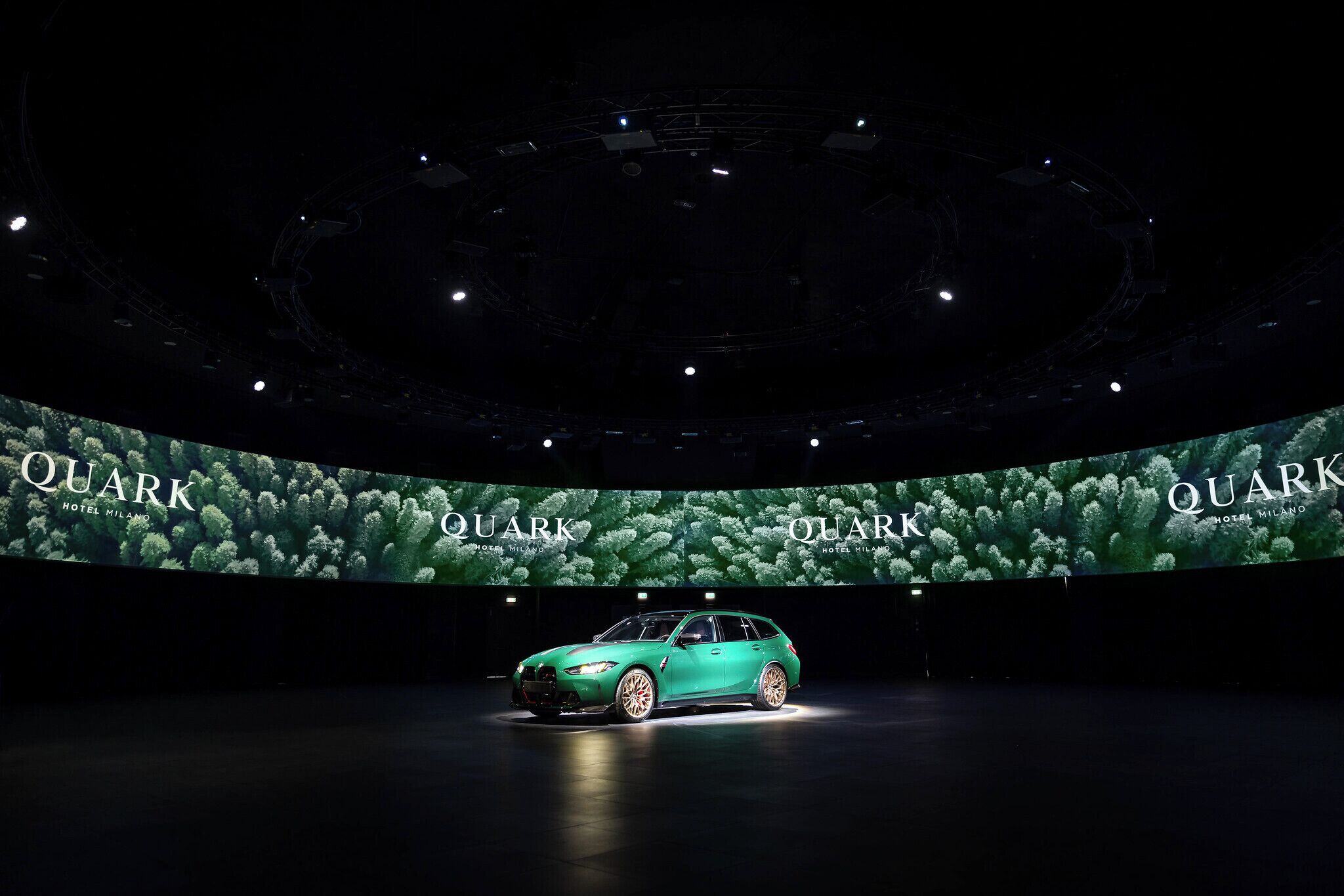 Um carro verde em exposição, com a marcação 'QUARK HOTEL MILANO' ao fundo.